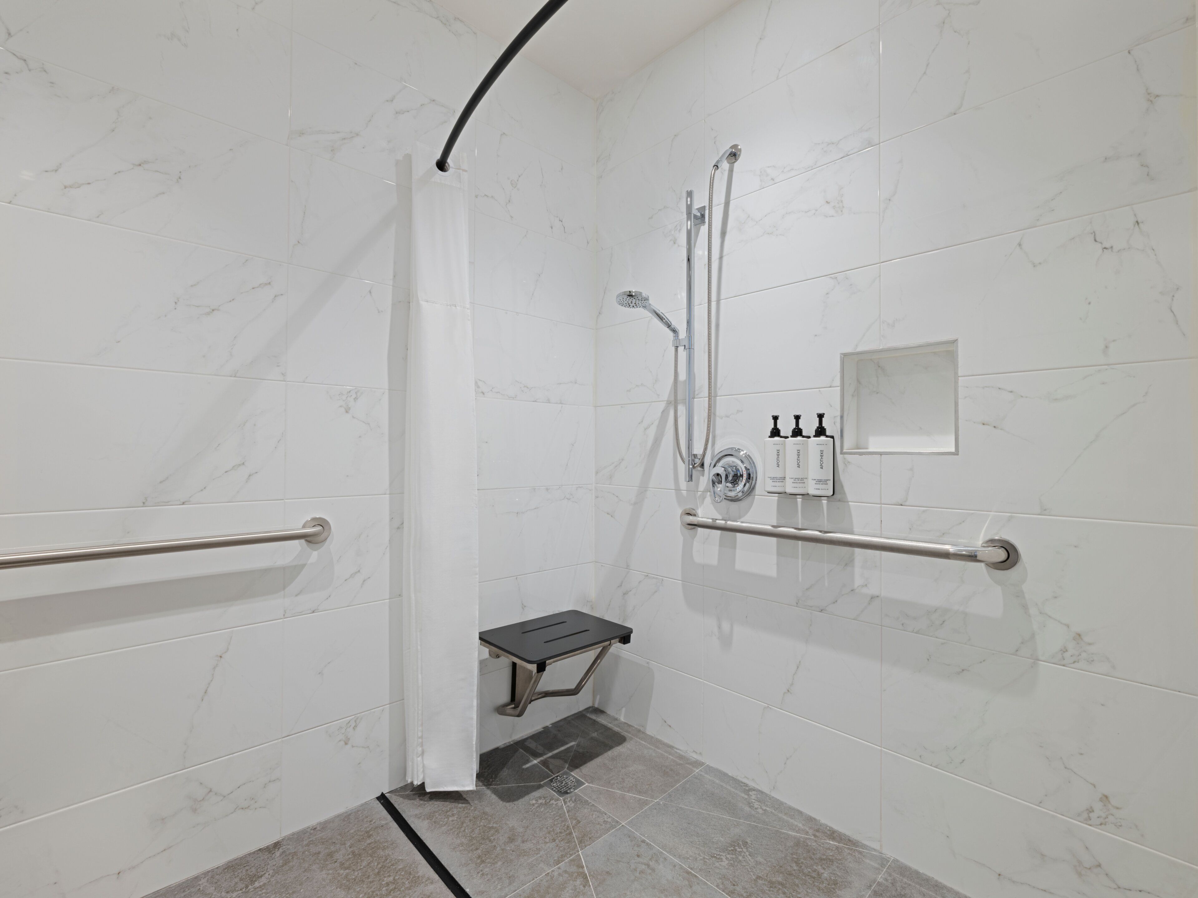 Casa de banho com azulejos de mármore branco, chuveiro com chuveiro manual, barra de apoio e um pequeno armário com artigos de higiene pessoal.