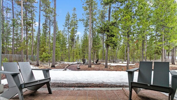 Um pátio com duas cadeiras Adirondack e uma mesa de vidro, rodeado por uma floresta de pinheiros altos e um chão coberto de neve.