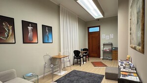 Uma sala de estar moderna com um sofá, uma mesa de centro e um tapete. Há três obras de arte emolduradas na parede, uma porta de madeira e uma mesinha com revistas.