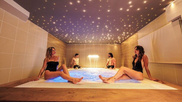 Uma banheira de hidromassagem com quatro pessoas sentadas ao redor, cercada por paredes azulejadas e um teto com pequenas luzes.