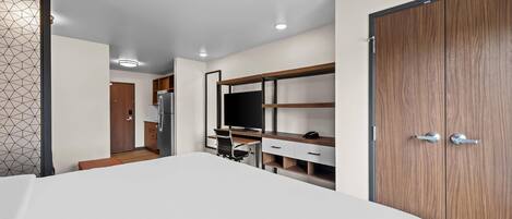 Quarto de hotel moderno com uma cama grande, uma escrivaninha com cadeira, um guarda-roupa e uma cozinha compacta.