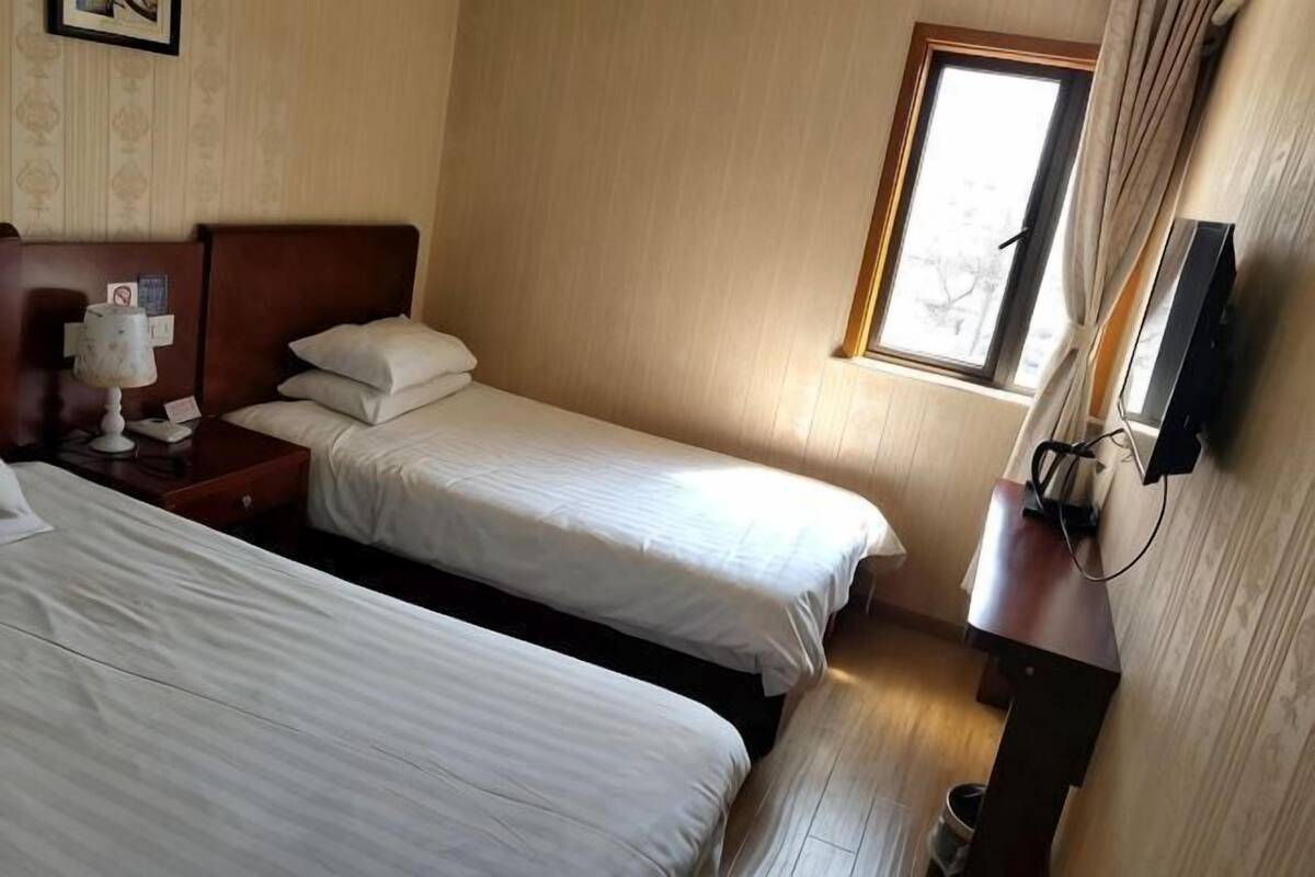 Quarto de hotel com duas camas, mesa de cabeceira, luminária, televisão e janela.