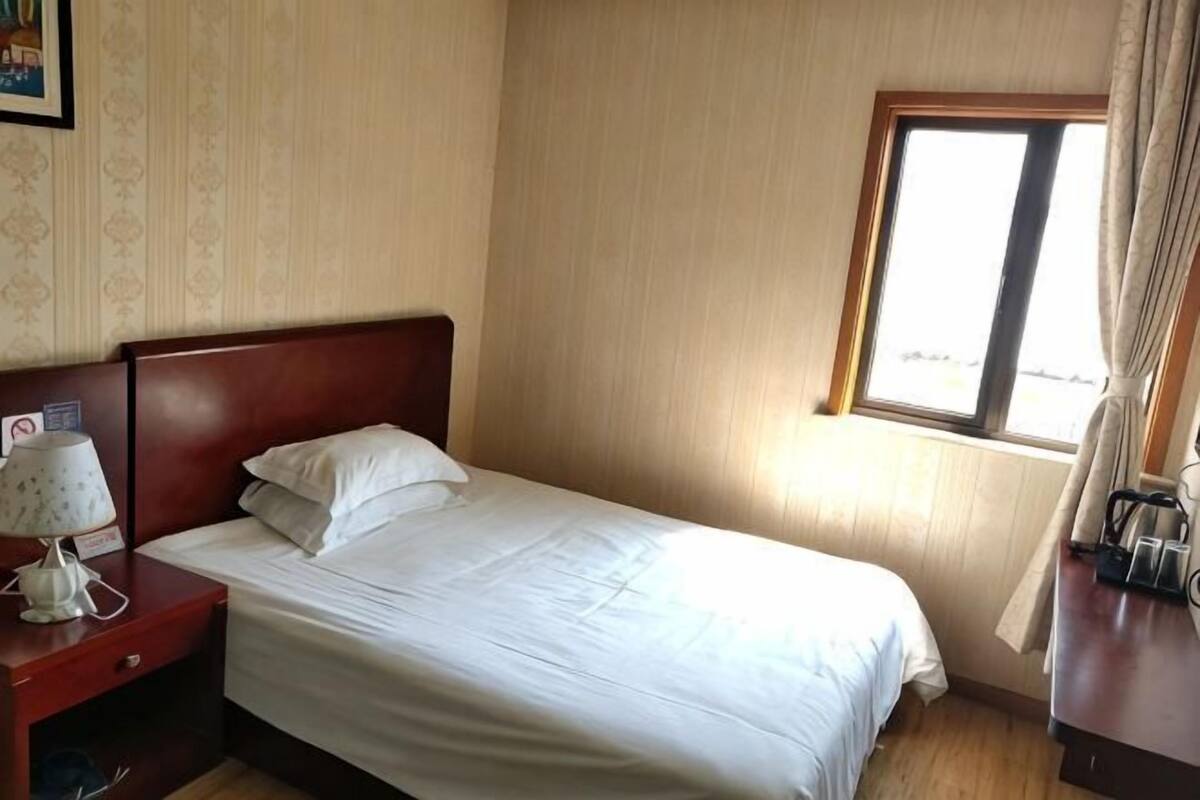 Uma cama de solteiro com cabeceira de madeira, uma mesa de cabeceira com abajur e uma janela com cortinas.