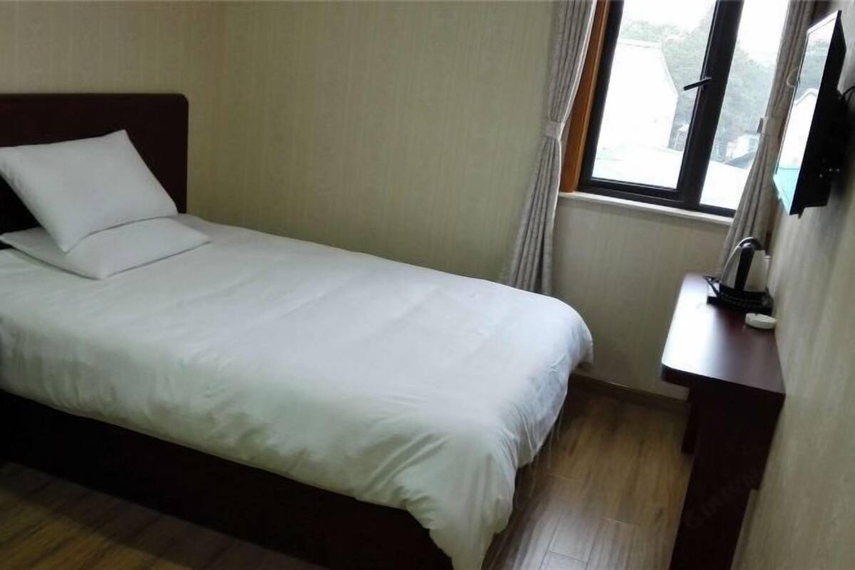 Uma cama de solteiro com roupa de cama branca, cabeceira de madeira, mesa de cabeceira com uma luminária e uma janela com cortinas.