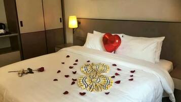Quarto de hotel com cama bem arrumada, um balão em formato de coração e pétalas de rosas dispostas sobre a cama.