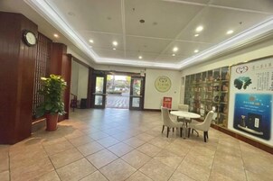 Um saguão de hotel com balcão de recepção, uma planta em vaso, área de estar e uma parede com cartazes informativos.