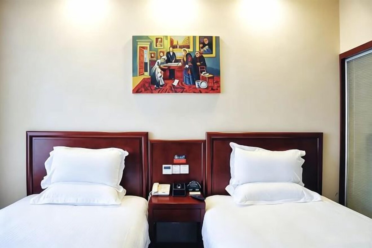 Quarto de hotel com duas camas, um quadro na parede, uma mesinha de cabeceira com telefone e relógio, e uma janela com persianas.