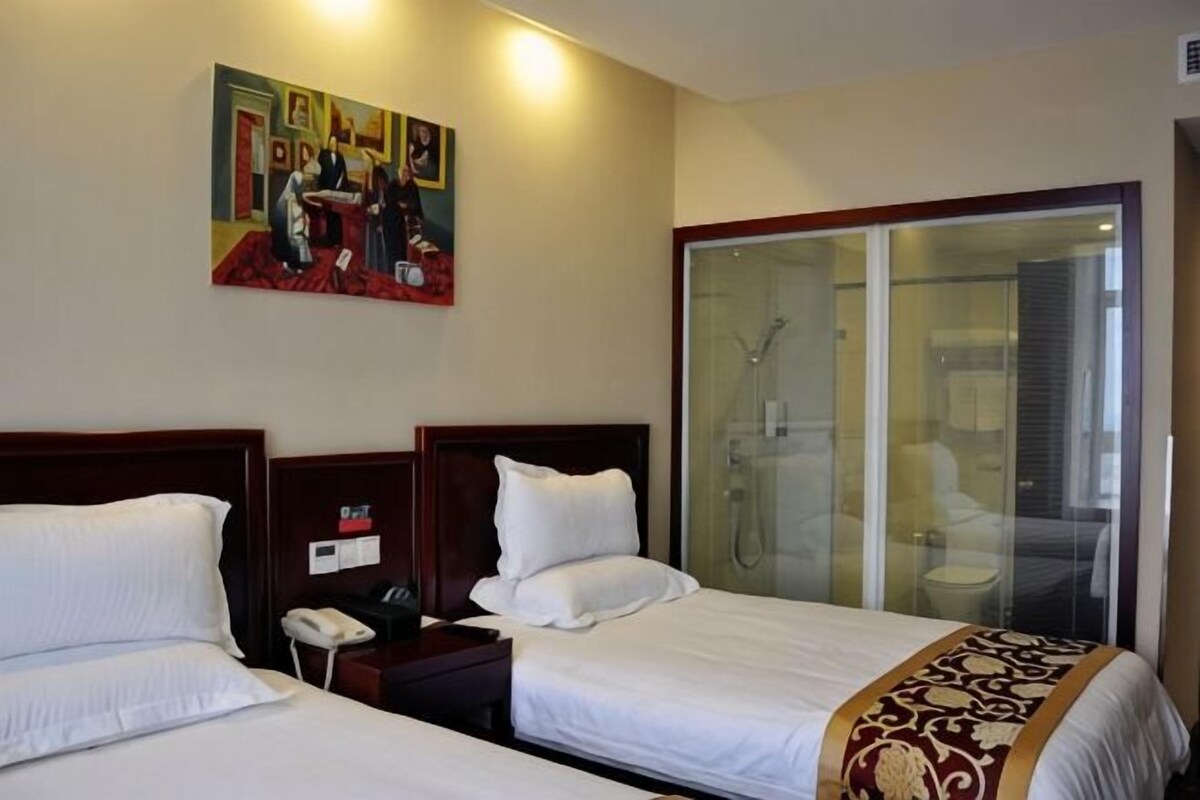 Quarto de hotel com duas camas, um quadro na parede, um telefone na mesa de cabeceira e zona de duche com divisória de vidro.