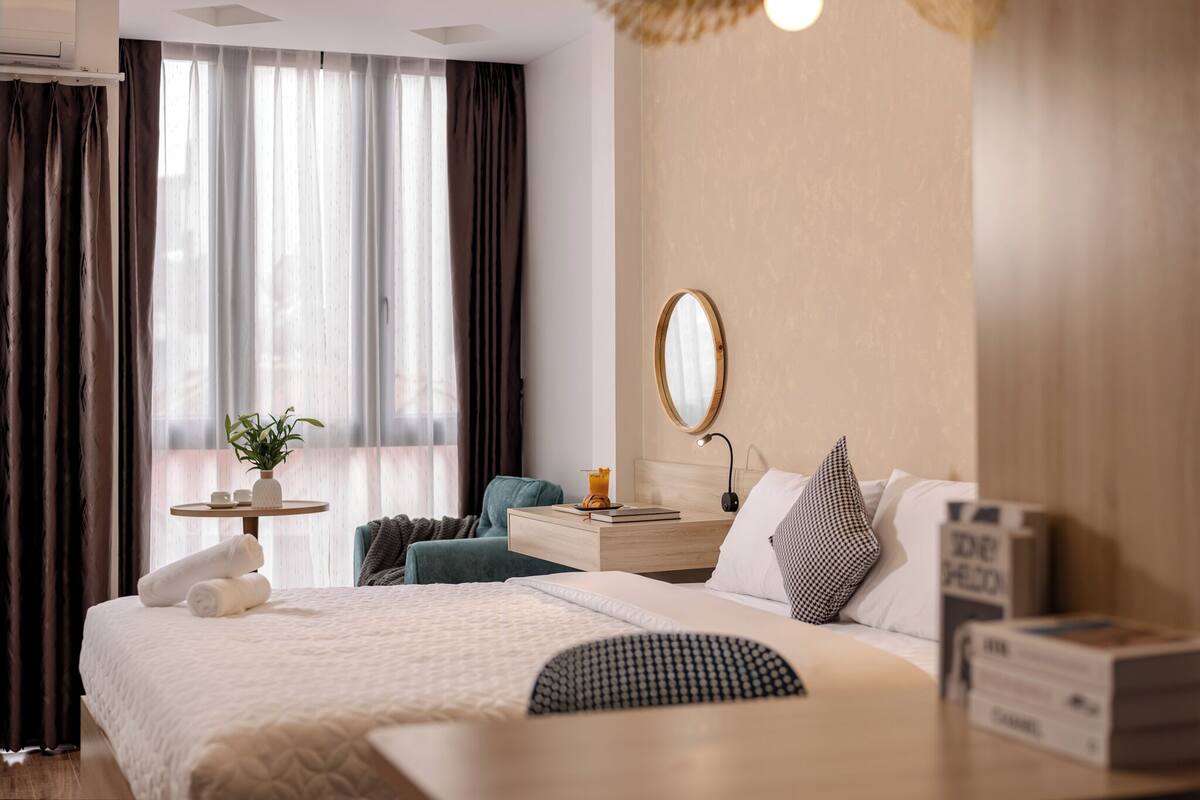 Quarto de hotel moderno com uma cama grande, mesa de cabeceira com luminária, espelho e vista para o exterior através de cortinas translúcidas.