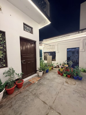 Um pátio com diversos vasos de plantas, uma porta marrom e uma parede branca com detalhes decorativos.
