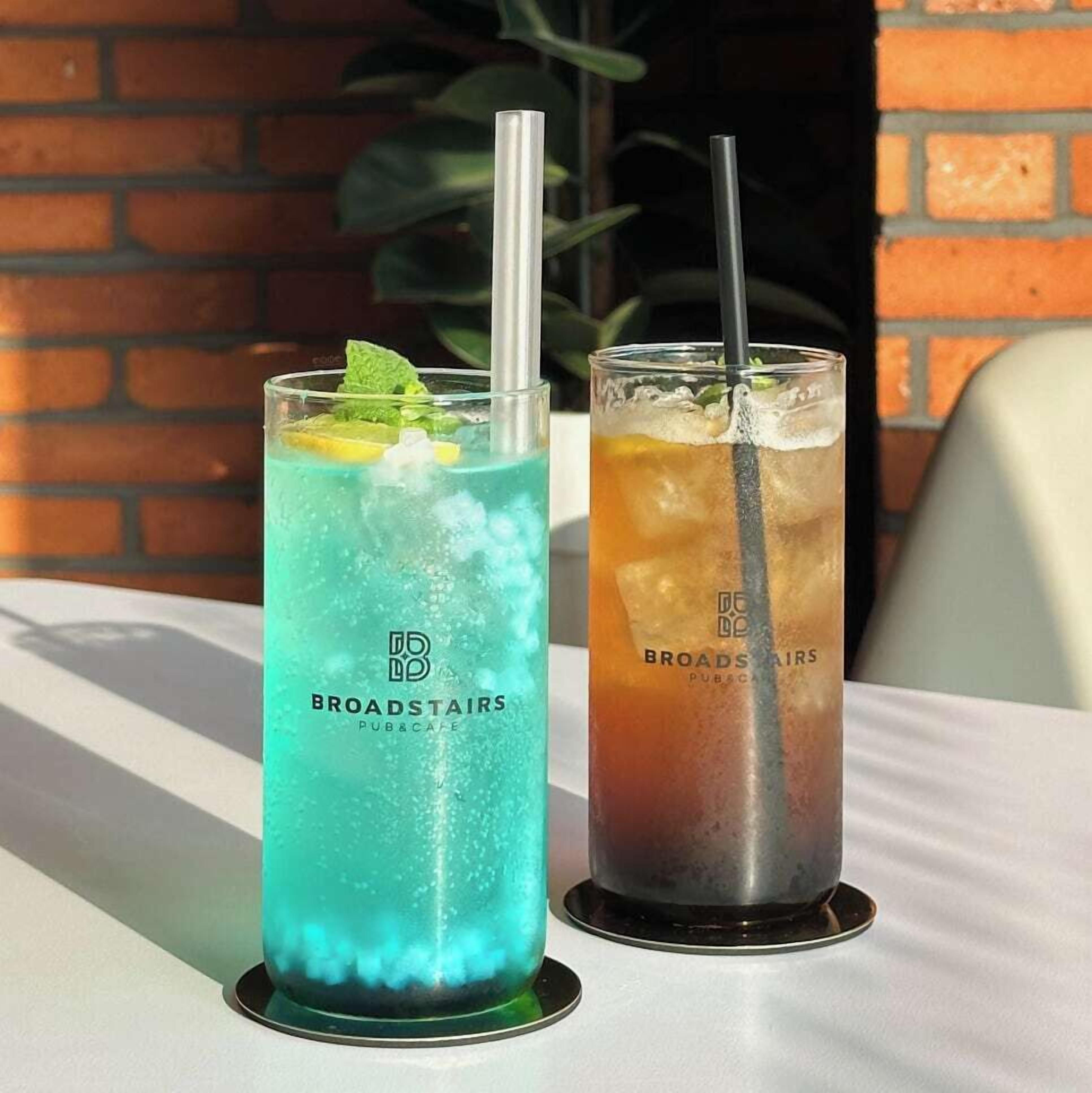 Duas bebidas sobre uma mesa, com um muro de tijolos e uma planta ao fundo.