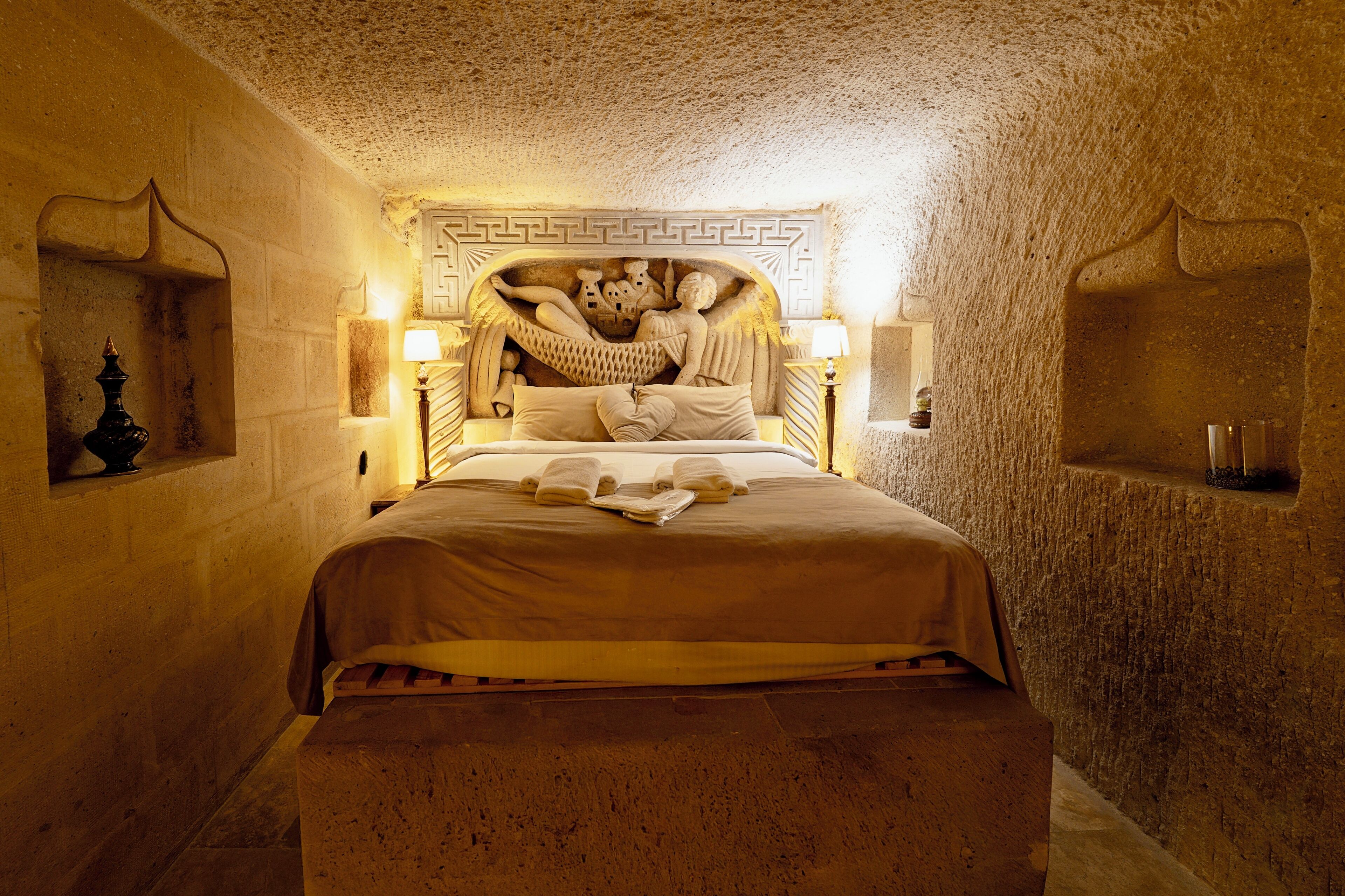 Quarto com paredes de pedra, uma cama, duas luminárias fixadas na parede e uma pequena prateleira com velas.