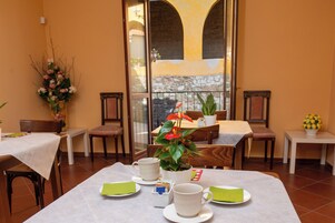 Uma área de jantar com mesas preparadas para dois, cada uma com toalha de mesa branca, xícara, pires e guardanapo. Há uma planta em vaso com uma flor vermelha em uma das mesas.