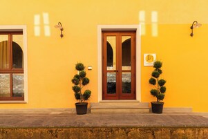 Um edifício com fachada amarela, duas plantas topiárias em vasos e uma porta dupla com painéis de vidro.