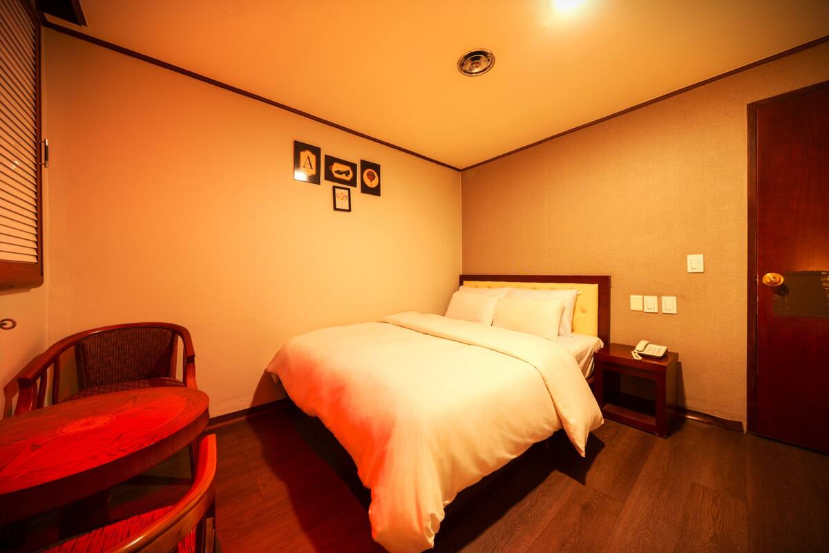Quarto de hotel com uma cama, mesa de cabeceira com telefone, uma cadeira e uma pequena mesa.
