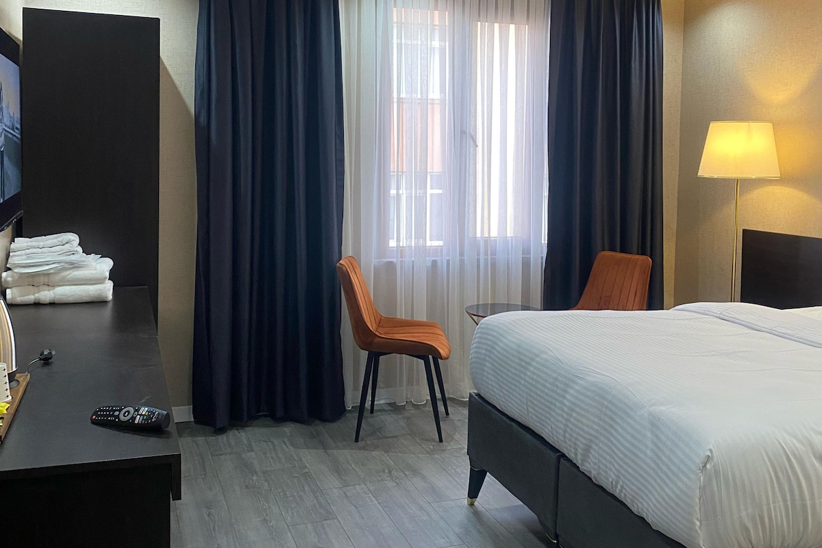 Quarto de hotel com uma cama, uma mesa com um controle remoto, uma cadeira, um abajur e uma televisão sobre um suporte.