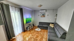 Uma sala de estar moderna com piso de madeira em padrão herringbone, um sofá seccional cinza, uma mesa de centro branca e uma TV de tela plana fixada na parede.