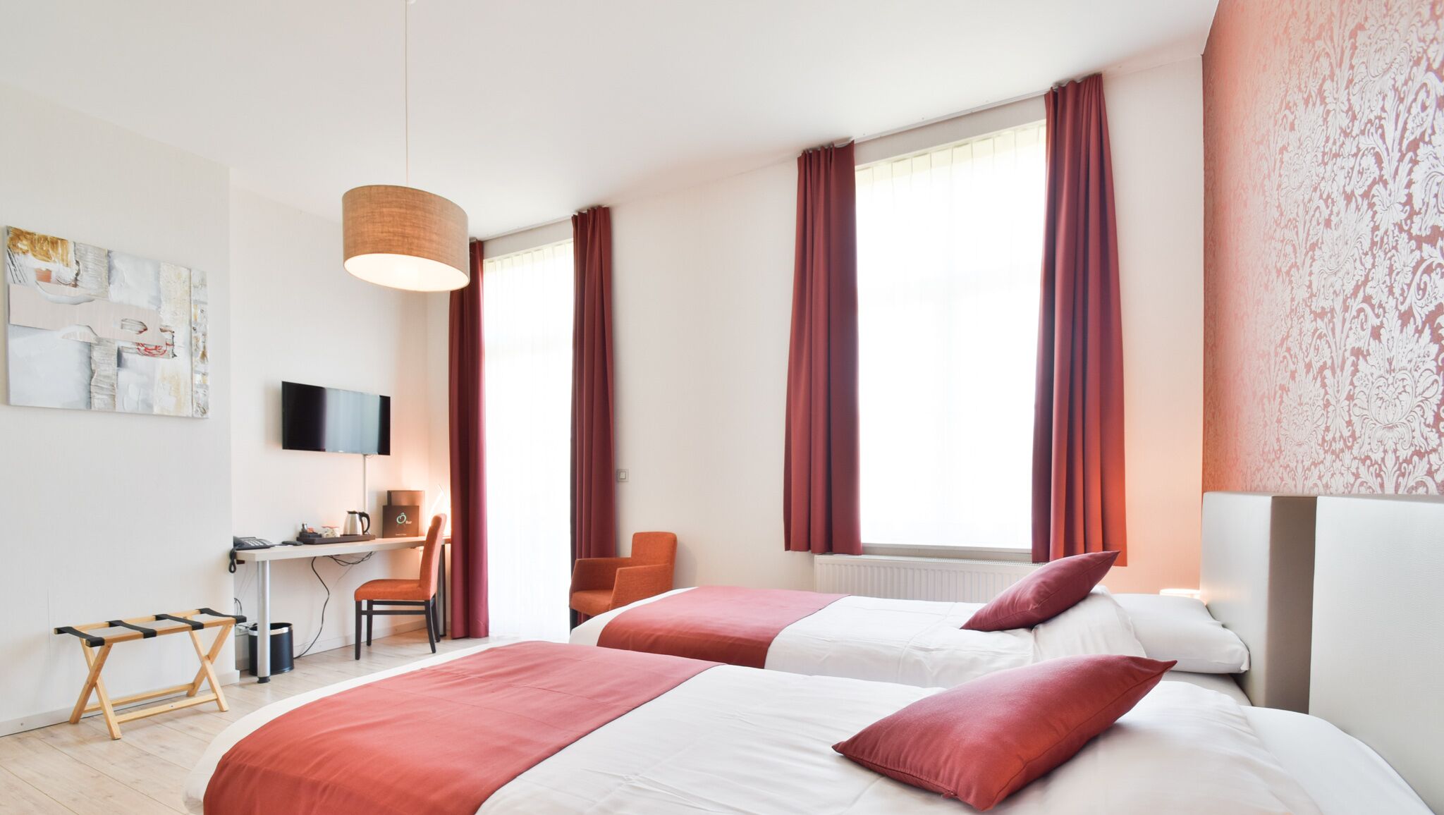 Quarto de hotel moderno com duas camas, uma escrivaninha com cadeira, televisão e uma janela grande com cortinas.