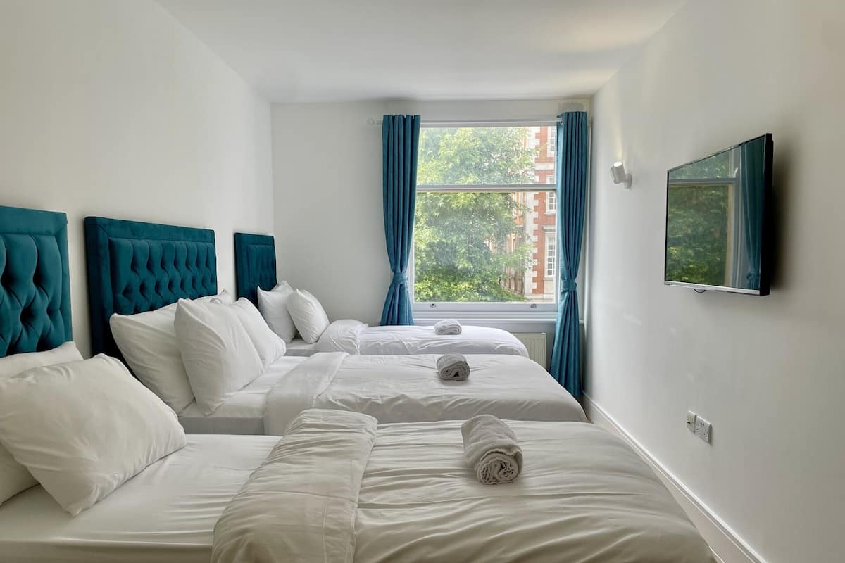 Quarto de hotel com duas camas, uma janela grande com cortinas, uma televisão de tela plana na parede e vista para o verde lá fora.