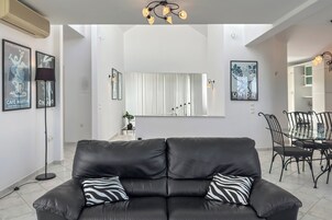 Uma sala de estar moderna com um sofá de couro preto, almofadas estampadas com padrão de zebra, uma área de jantar com mesa de vidro e cadeiras de metal preto, e um espelho grande acima do sofá.