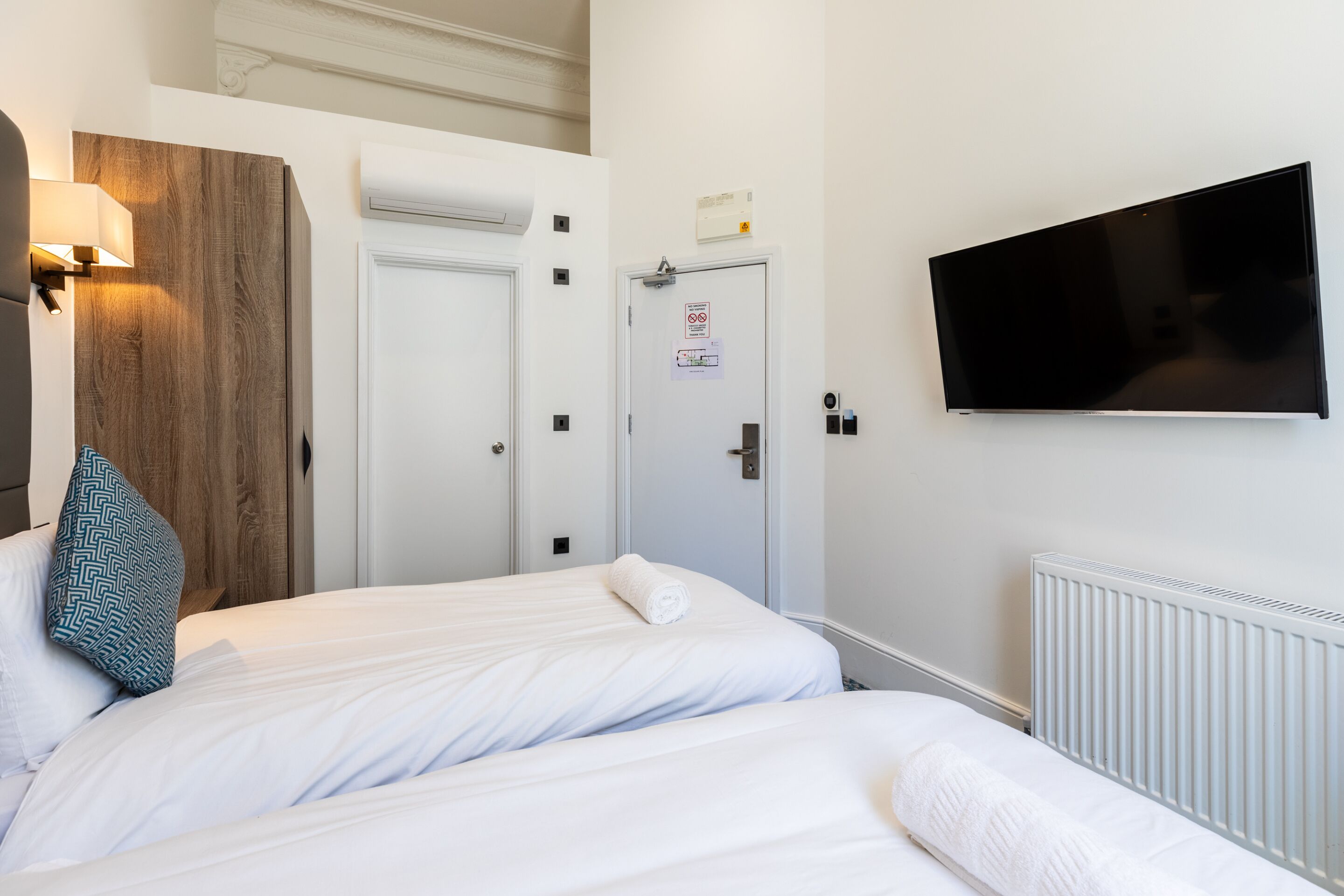 Quarto de hotel com duas camas, televisão de tela plana, cabeceira de madeira, porta branca e radiador.