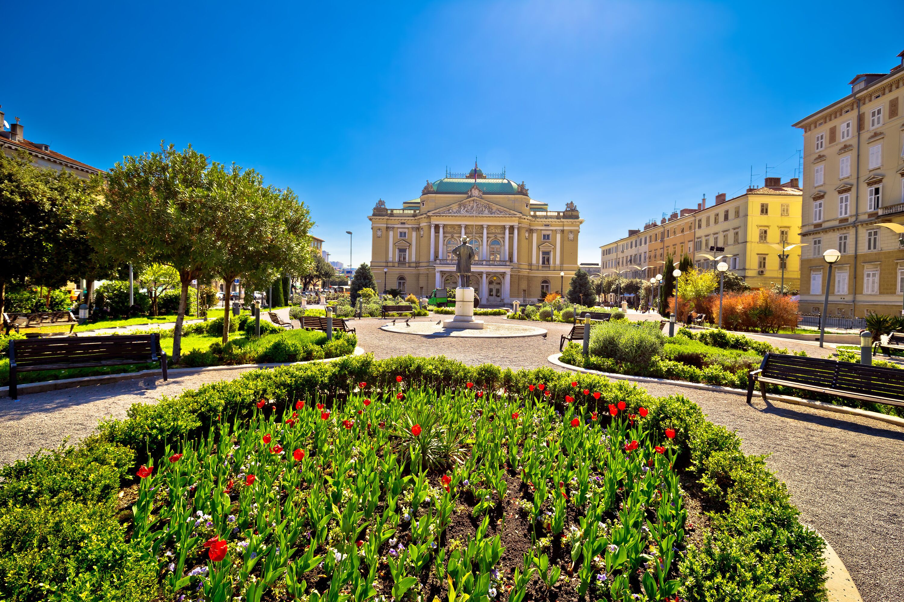 Uma praça com um monumento central, rodeada por bancos e um jardim florido.