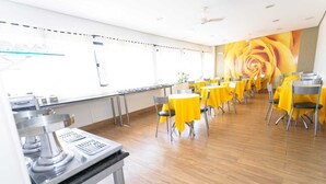 Uma área de refeições luminosa com mesas e cadeiras amarelas, uma janela grande com persianas e uma parede com um mural floral.