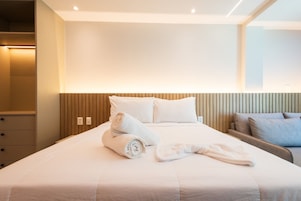 Quarto de hotel com uma cama bem arrumada, cabeceira de madeira e um espaço de estar com uma cadeira e uma mesa pequena.