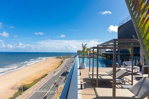 Varanda com vista para a praia e oceano, cadeiras de descanso e área de piscina.