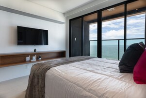Quarto moderno com cama grande, televisão de tela plana na parede, mesa de consola de madeira e vista para o mar através de amplas janelas.