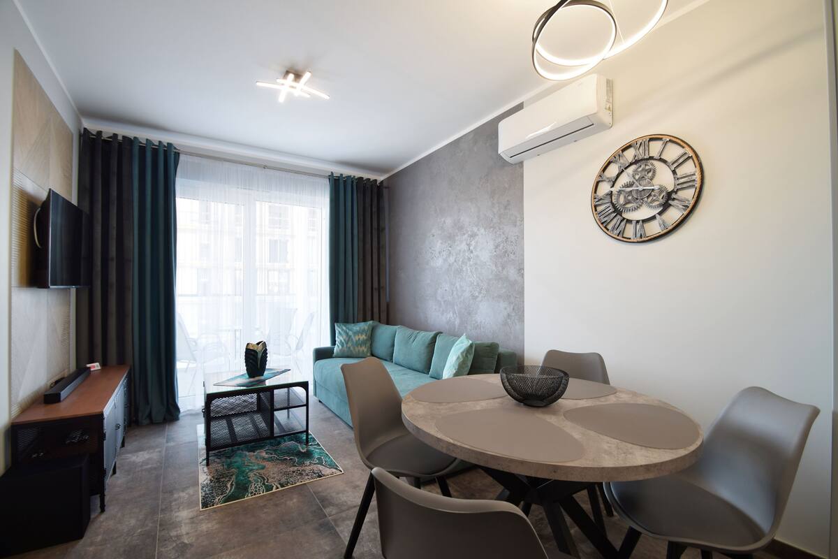 Uma sala de estar moderna com uma mesa de jantar redonda, cadeiras cinzas, um sofá e um relógio fixado na parede.