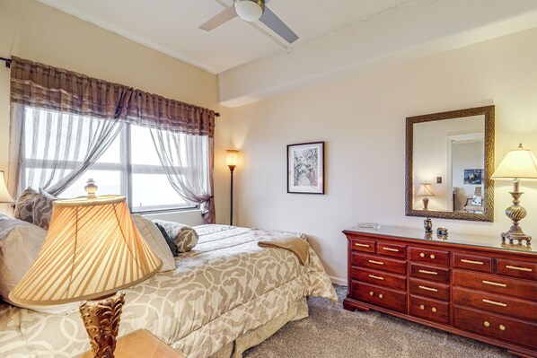 Quarto com cama, cômoda, espelho, abajur e janela com cortinas.