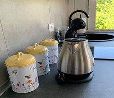 Uma bancada de cozinha com três recipientes brancos com tampas amarelas, uma chaleira de aço inoxidável e uma cafeteira.
