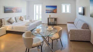 Quarto de hotel moderno com uma cama, uma mesa de jantar de vidro com cadeiras, um sofá e duas janelas.