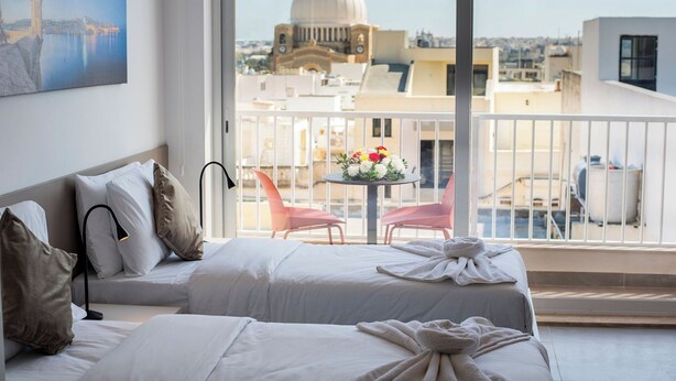 Quarto de hotel com varanda, cama com roupa de cama branca, uma pequena mesa com flores e um quadro na parede.