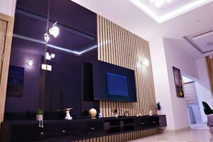 Uma sala moderna com uma televisão de tela plana grande fixada em uma parede de painéis de madeira, um rack de entretenimento escuro abaixo e um lustre no teto.