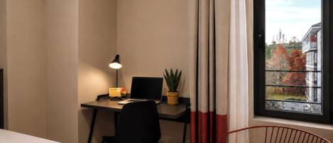 Quarto de hotel com cama, escrivaninha com laptop, cadeira, planta em vaso e janela com cortinas.