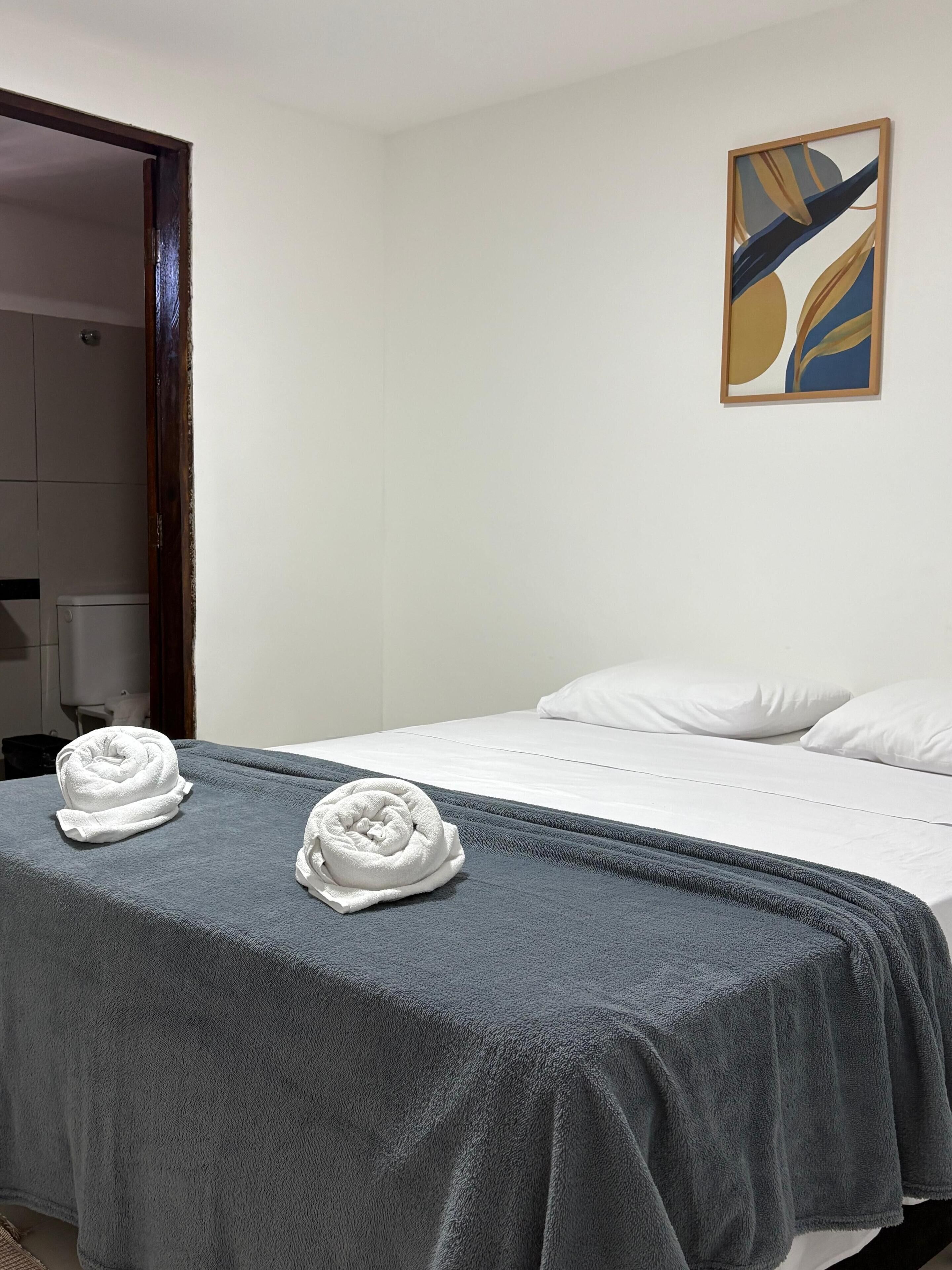 Quarto de hotel com cama, duas toalhas enroladas, um cobertor cinza e um quadro na parede.