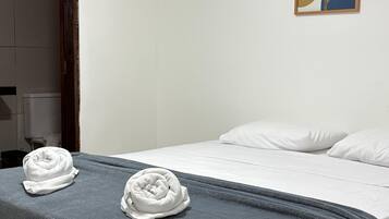 Quarto de hotel com cama, duas toalhas enroladas, um cobertor cinza e um quadro na parede.