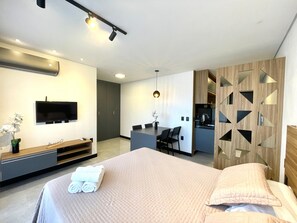 Quarto de hotel moderno com cama grande, escrivaninha, televisão e cozinha compacta.