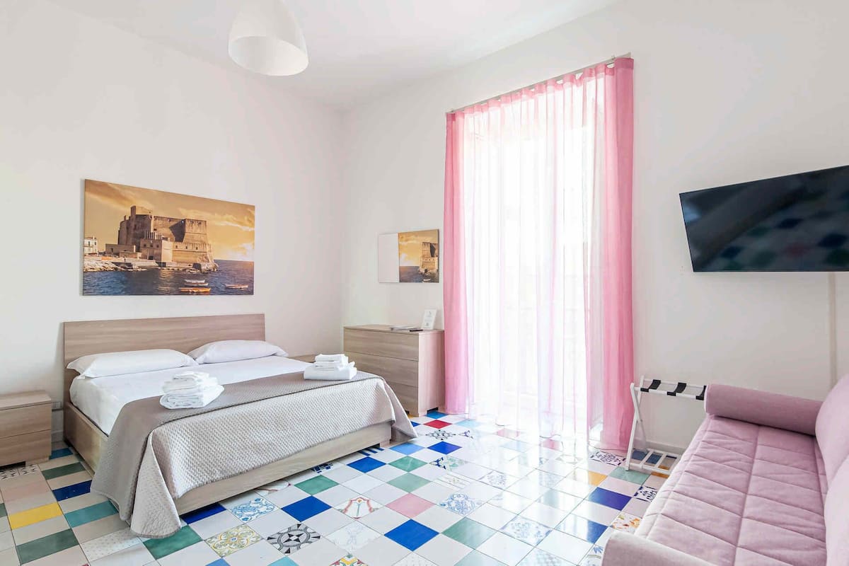 Quarto com cama, mesas de cabeceira, televisão e um sofá rosa.