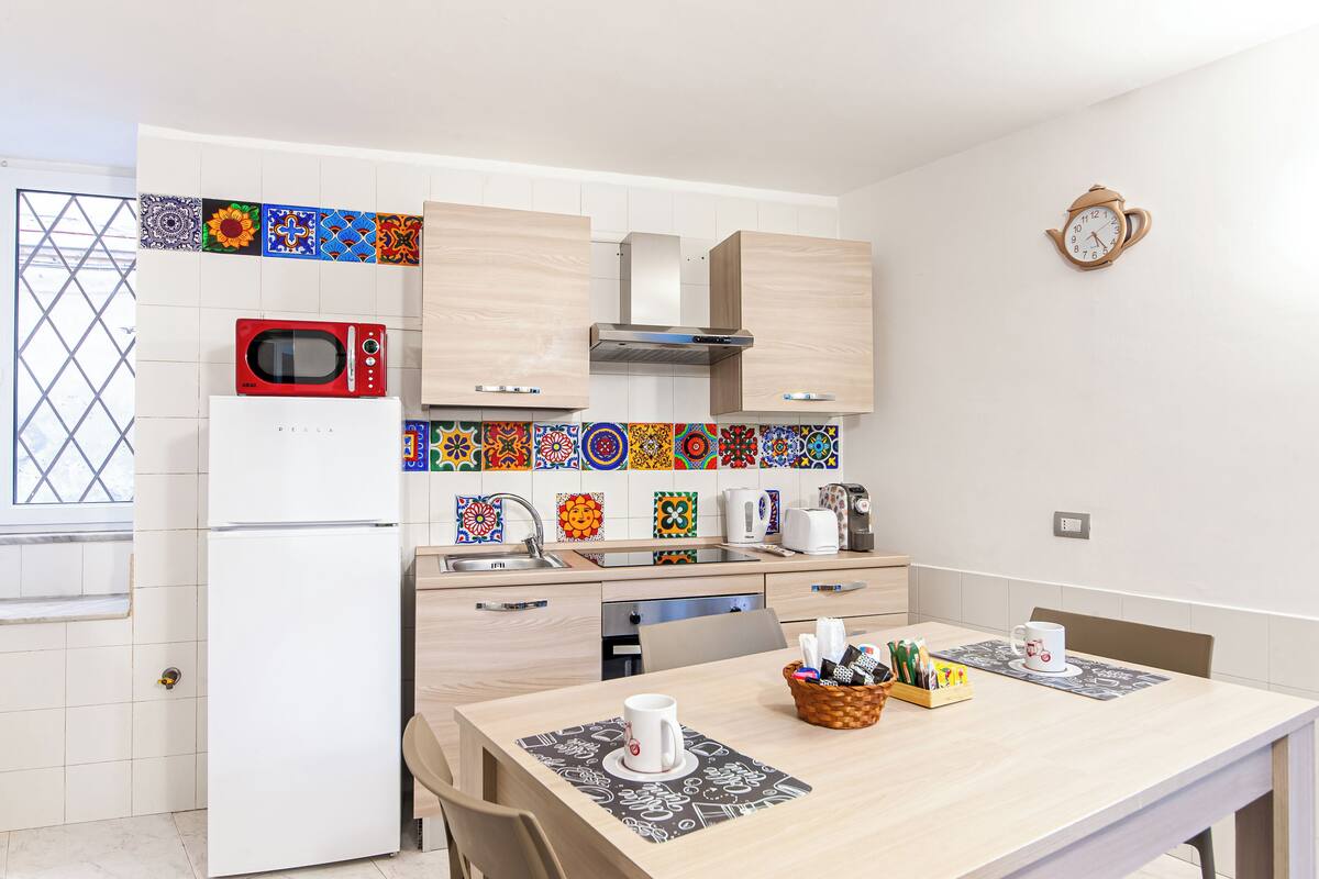 Cozinha com micro-ondas, fogão e pia, mesa de jantar com jogo americano e azulejos coloridos na parede.