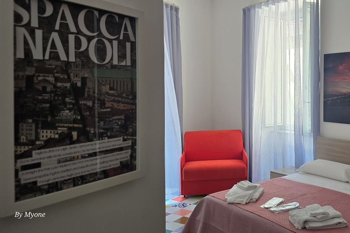 Quarto de hotel com uma cama, uma cadeira vermelha, uma janela com cortinas, um cartaz de paisagem urbana e uma pintura de pôr do sol.