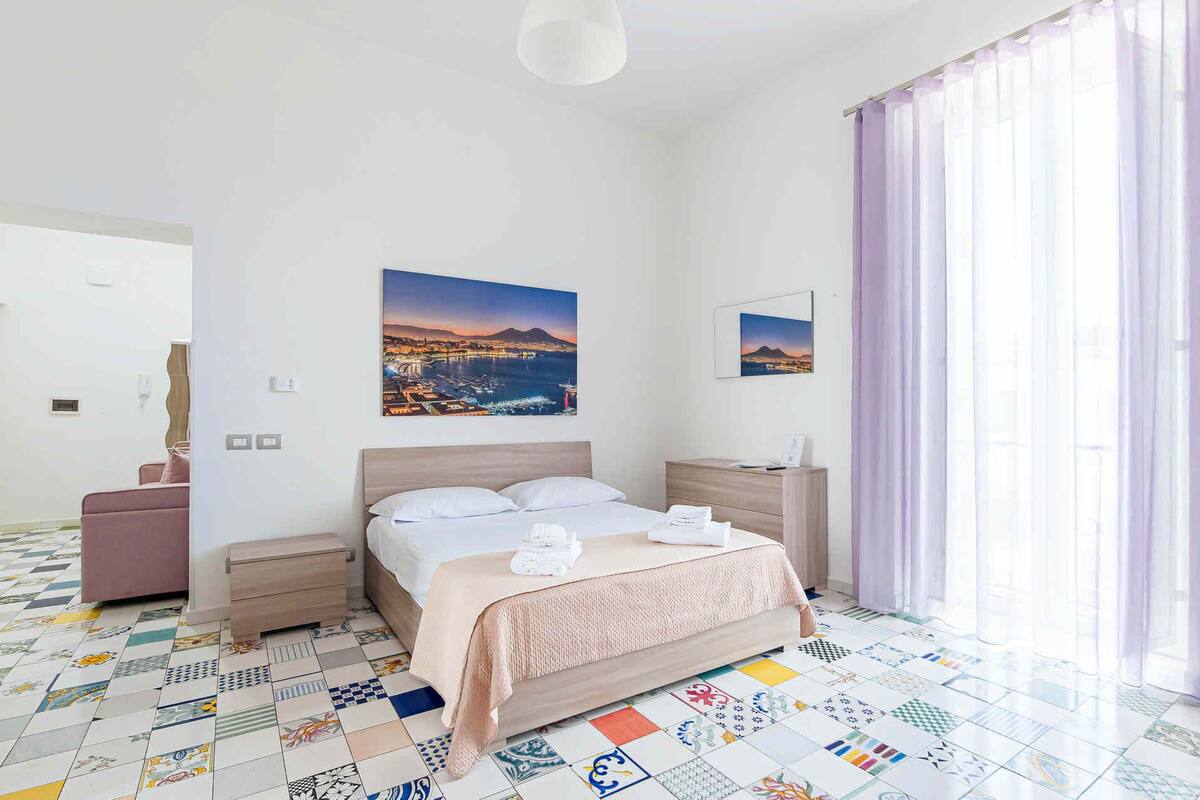 Quarto de hotel com cama, mesa de cabeceira, cômoda e janela com cortinas.