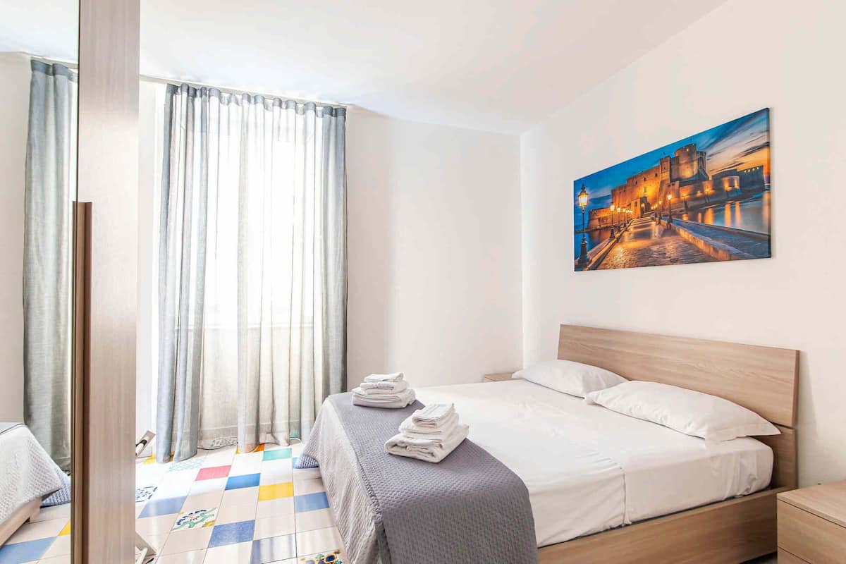 Quarto de hotel com cama, pintura de uma paisagem urbana, janela com cortinas e piso de azulejos.