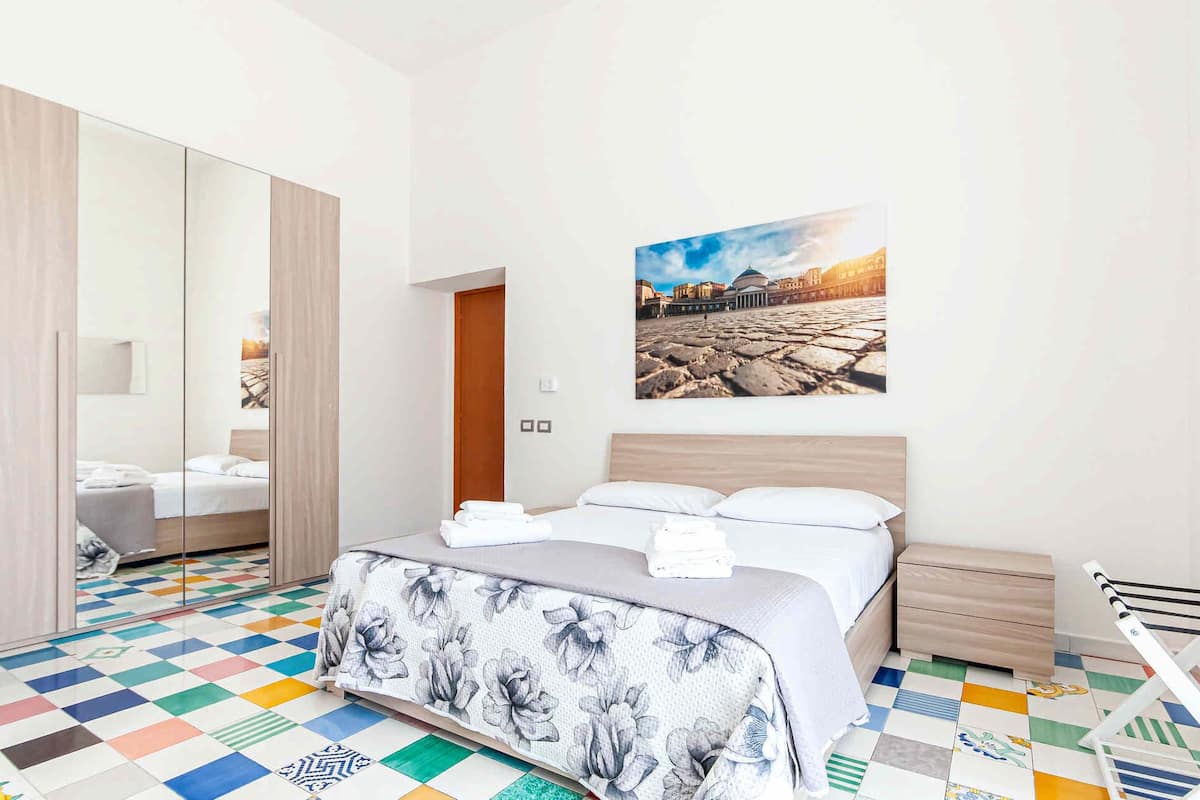 Quarto de hotel com cama, espelho, guarda-roupa, mesa de cabeceira e quadro pendurado na parede.