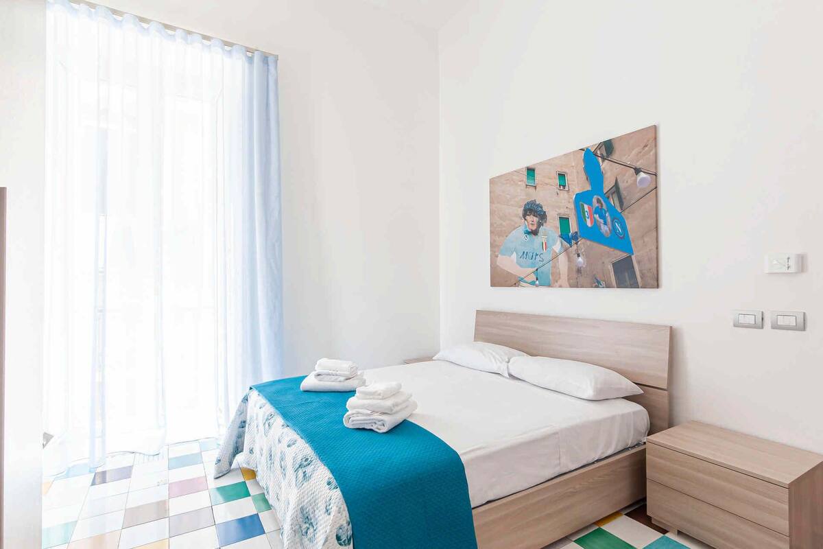 Quarto de hotel com cama, mesa de cabeceira, quadro na parede e janela com cortinas.
