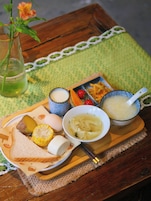 Uma mesa de madeira com toalha de mesa verde, um prato de comida incluindo pão, milho, ovos e sopa, e um pequeno vaso com flores.