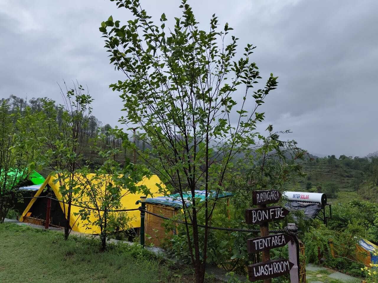 Um camping com uma tenda amarela, um banheiro e placas indicando área para fogueira, área de tendas e área de recreação.