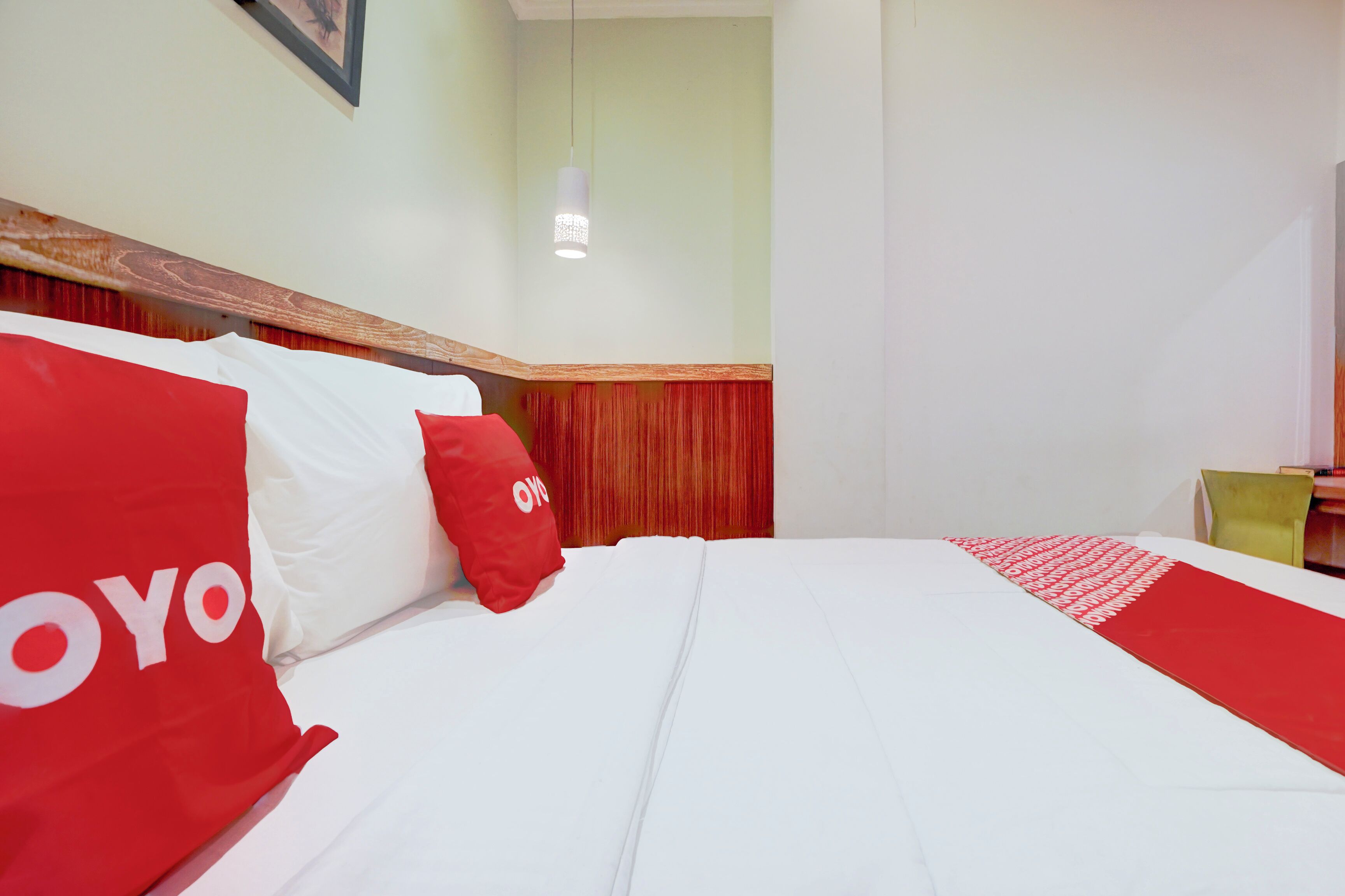 Quarto de hotel com cama, almofadas vermelhas e brancas, colcha vermelha e branca, cabeceira de madeira, luminária pendente e uma cadeira verde.